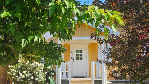 Uma casa com porta branca, varanda de madeira e pequeno jardim com vasos de plantas e flores.