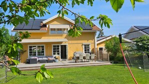Uma casa de dois andares com um amplo jardim frontal, uma área de refeições com móveis de exterior e uma quadra de tênis visível.