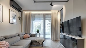Uma sala de estar moderna com um sofá cinzento, uma televisão de tela plana sobre um móvel, uma janela grande com cortinas translúcidas e uma pequena mesa lateral com uma planta.