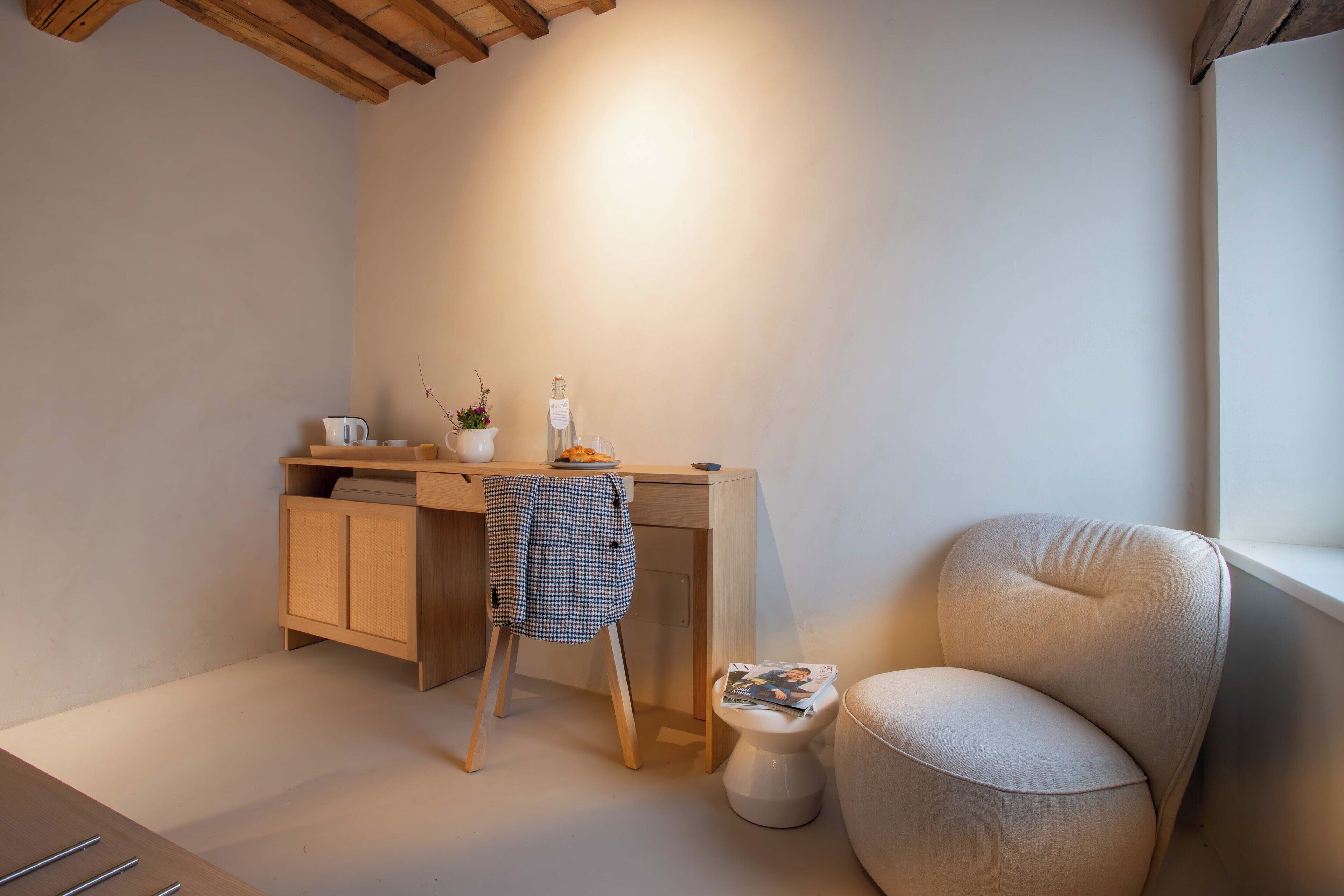 Quarto minimalista com escrivaninha de madeira, uma cadeira, um banquinho e uma janela.