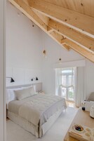 Quarto com uma cama grande, vigas de madeira no teto, duas luminárias pendentes, uma janela com cortinas e uma pequena mesa com xícara e pires.