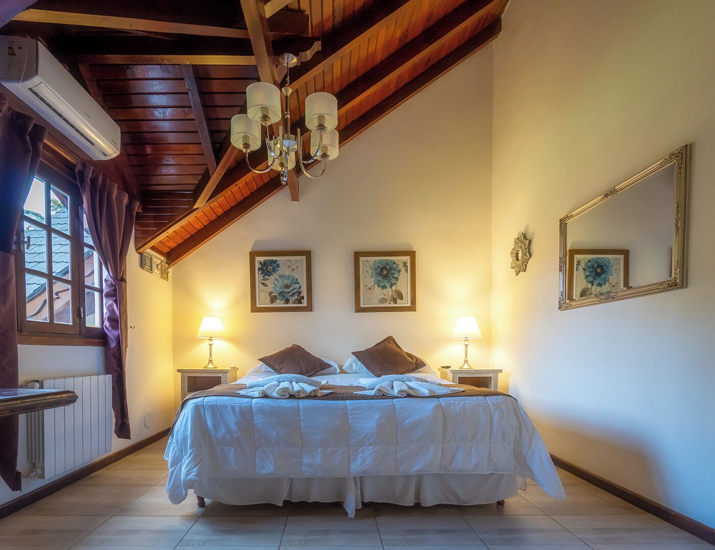 Quarto com teto de madeira, cama com roupa de cama branca, duas luminárias de cabeceira e arte floral nas paredes.