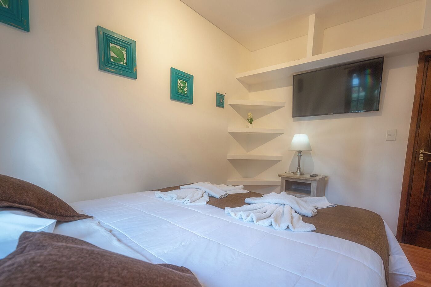 Quarto de hotel com uma cama, televisão na parede e uma mesinha com abajur.