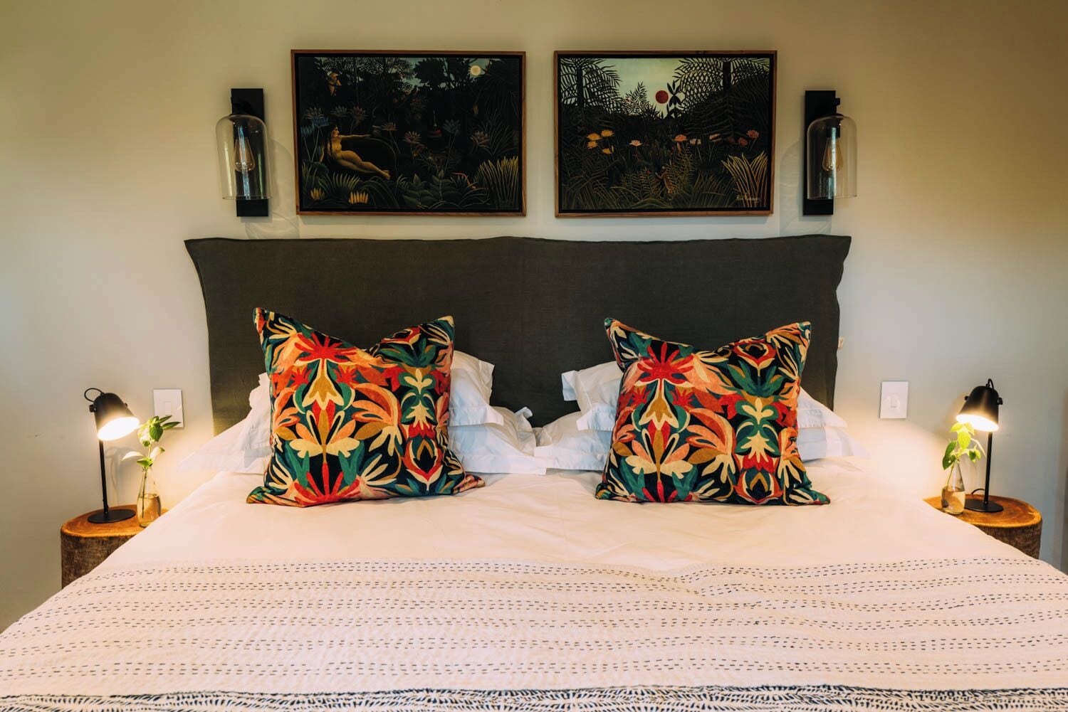 Cama bem arrumada com duas almofadas decorativas, cabeceira escura e duas luminárias fixadas na parede.