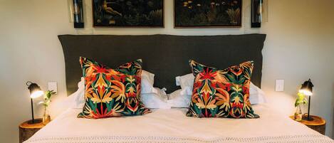Cama bem arrumada com duas almofadas decorativas, cabeceira escura e duas luminárias fixadas na parede.