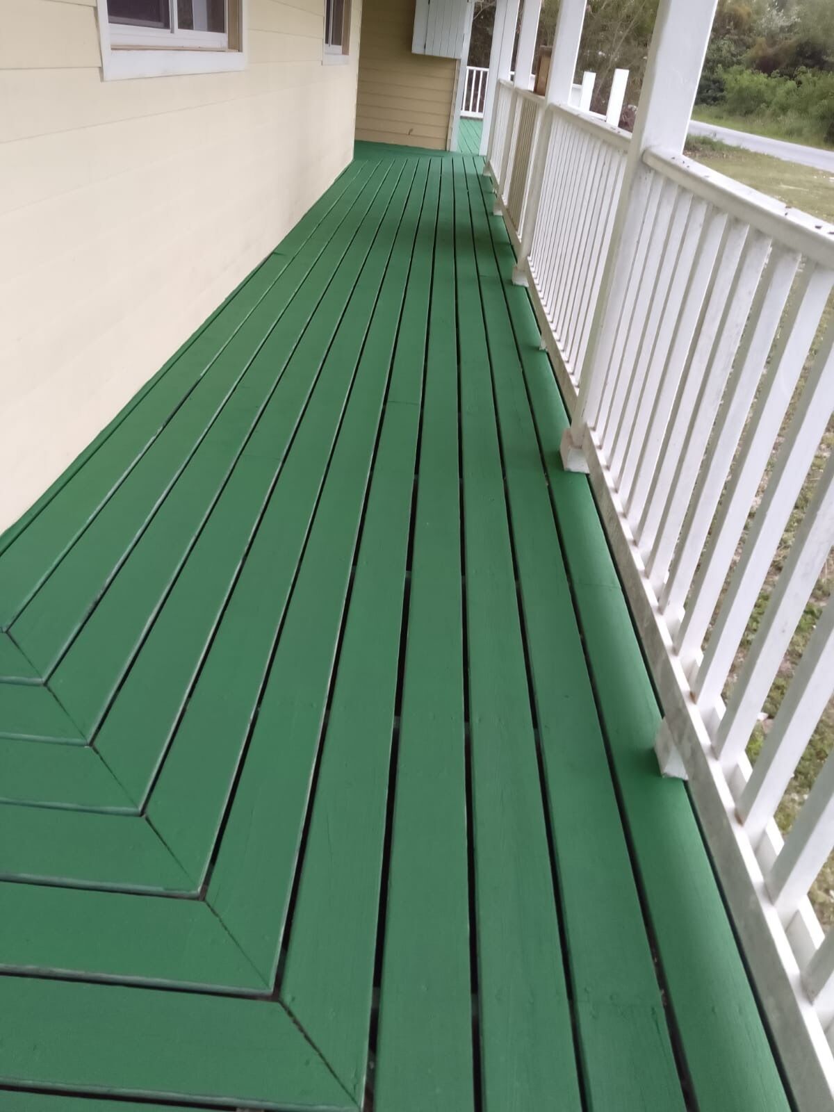 Um deck de madeira verde com guarda-corpos brancos, que dá acesso a um edifício bege com janela.