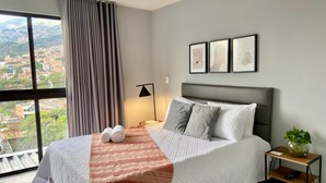 Quarto de hotel moderno com uma cama grande, vista para a paisagem urbana e uma pequena mesinha de apoio com uma planta.