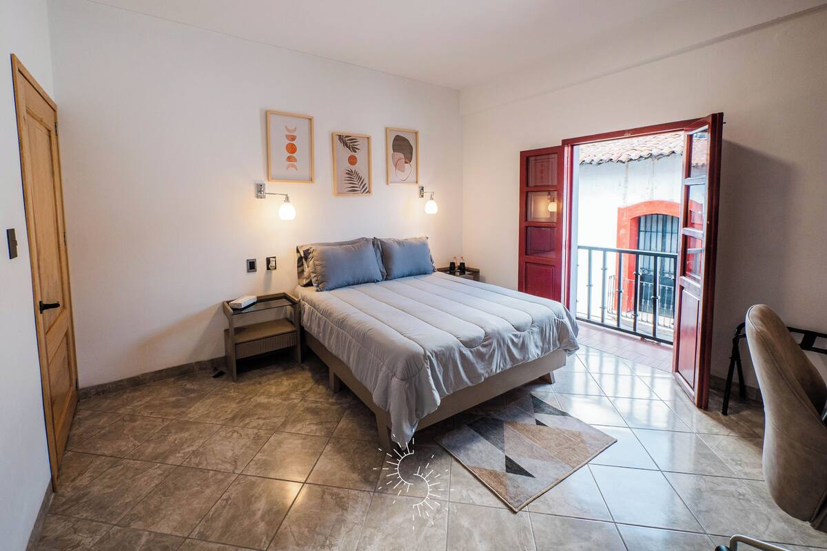 Quarto com cama, mesas de cabeceira, uma cadeira e vista para o exterior.