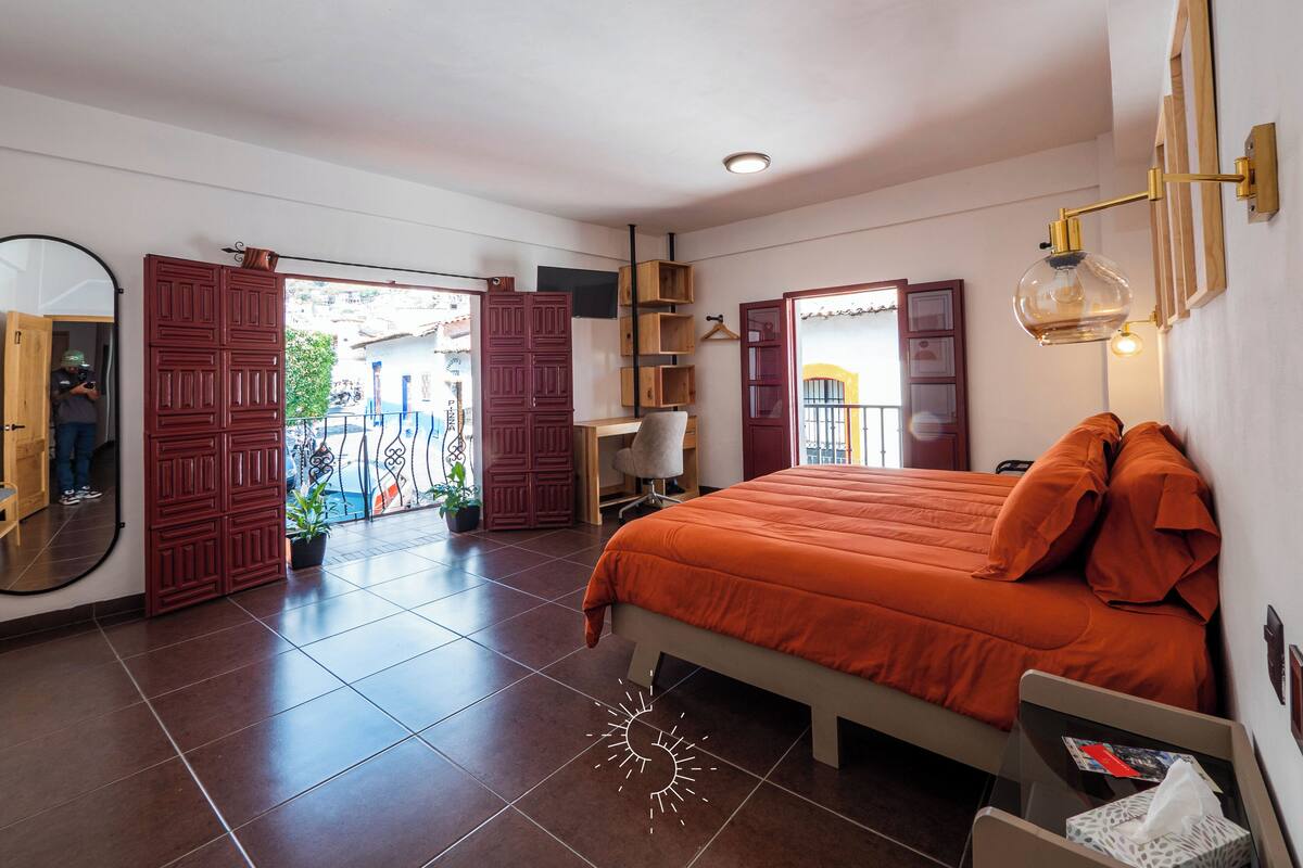 Quarto com cama, escrivaninha, espelho e vista para o exterior.