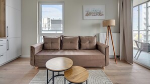Sala de estar moderna com um sofá bege, duas mesas redondas de madeira, um abajur e vista para a cidade através de grandes janelas.