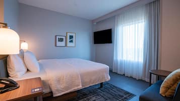 Quarto de hotel com uma cama, uma escrivaninha com abajur, uma televisão na parede e uma janela com cortinas.
