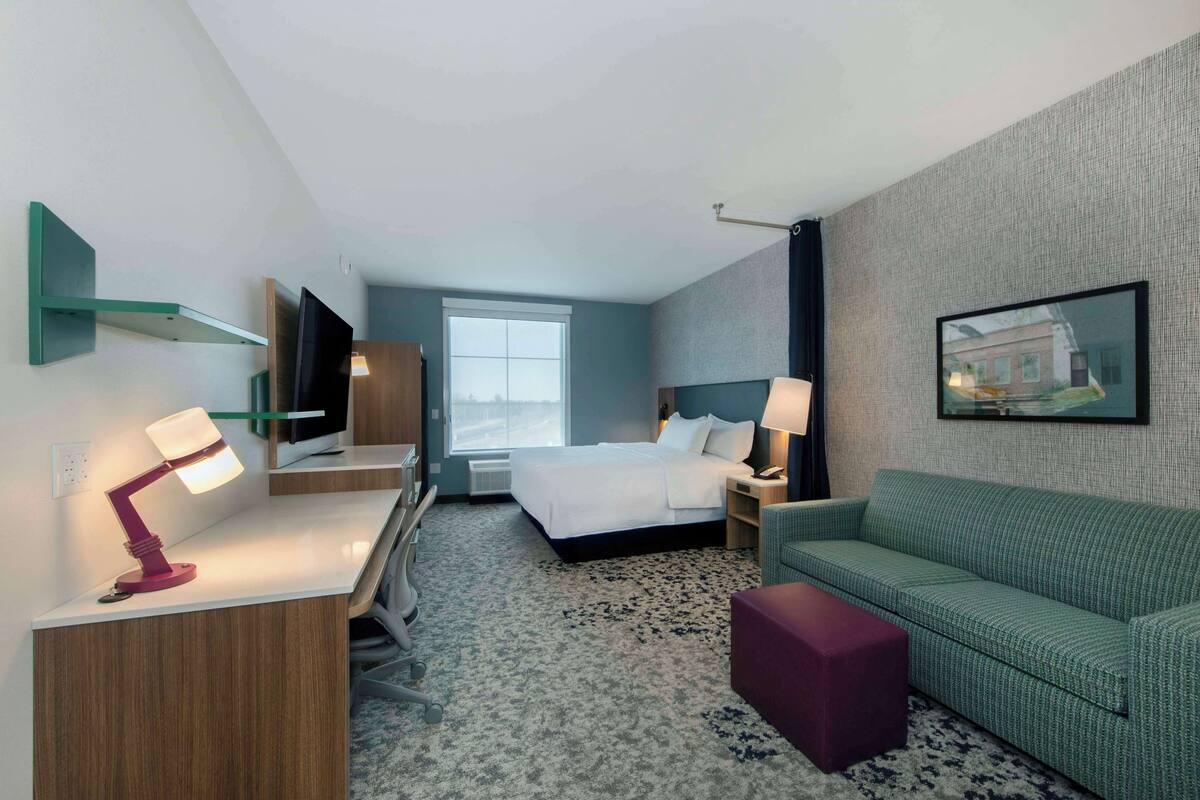 Quarto de hotel com cama, escrivaninha com abajur, sofá verde, mesa pequena e um quadro na parede.