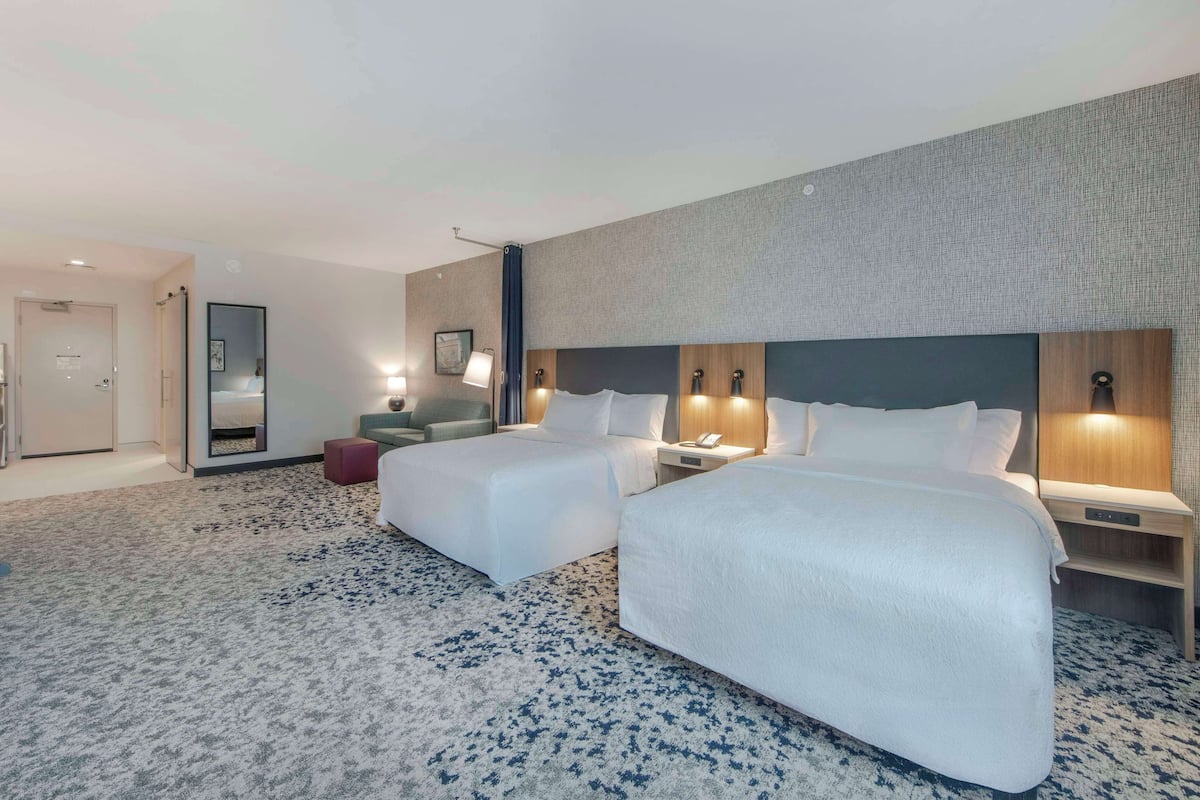 Quarto de hotel com duas camas, uma escrivaninha, uma cadeira e um pequeno espaço para sentar.