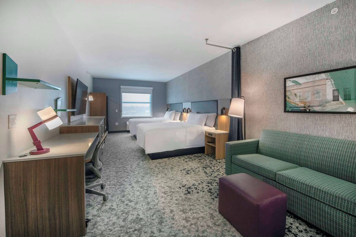 Quarto de hotel com cama, escrivaninha, sofá verde e televisão na parede.