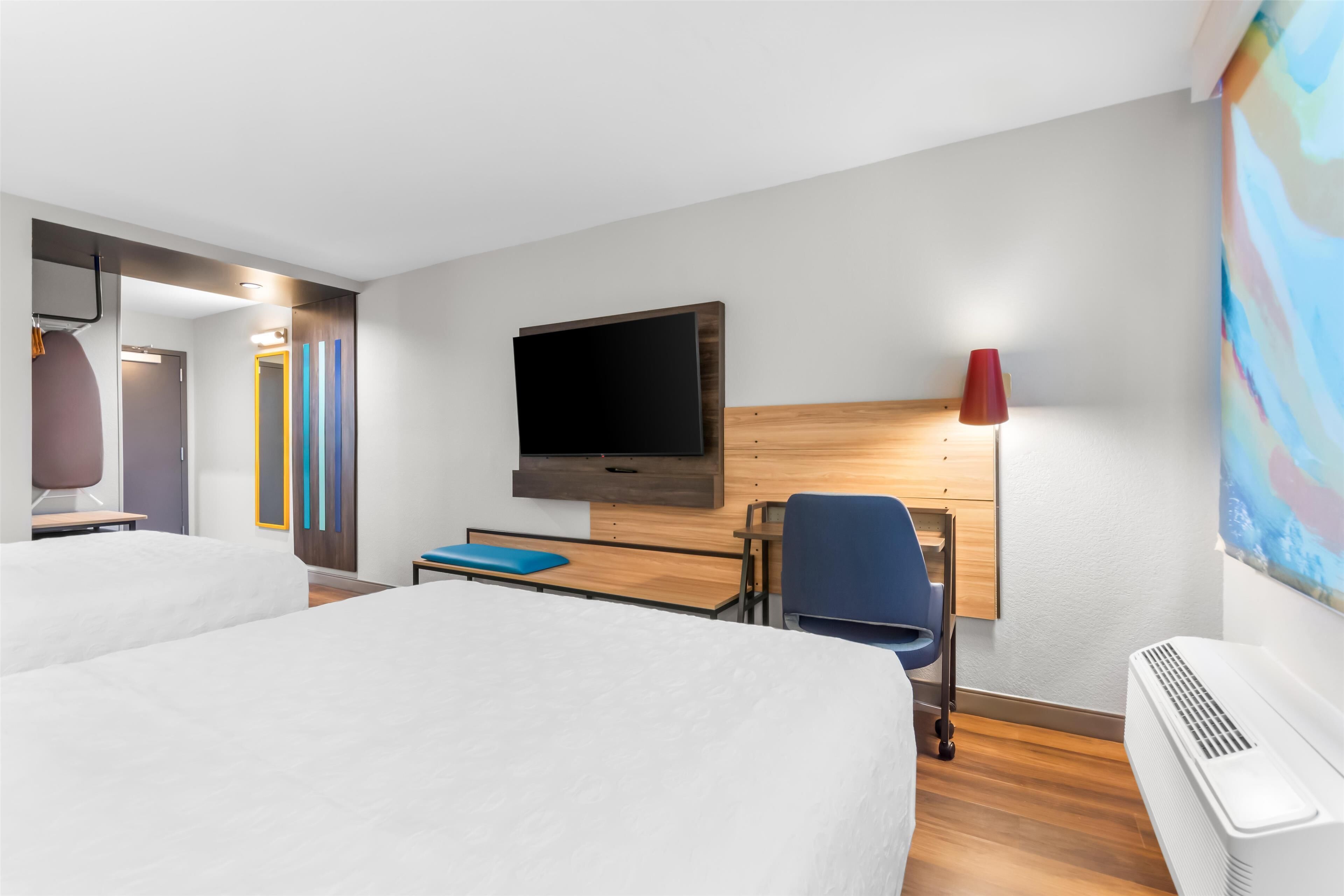 Quarto de hotel com duas camas, uma TV de tela plana, uma escrivaninha com cadeira, um abajur vermelho e um aquecedor fixado na parede.