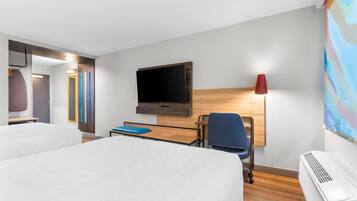 Quarto de hotel com duas camas, uma TV de tela plana, uma escrivaninha com cadeira, um abajur vermelho e um aquecedor fixado na parede.