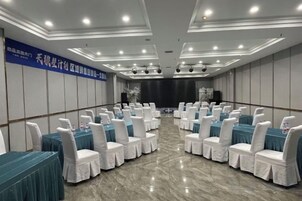 Uma sala de conferências com fileiras de mesas e cadeiras, um palco na frente e um banner na parede.