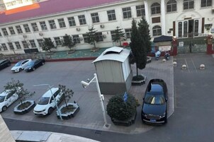Um estacionamento com vários carros, um pequeno edifício e um prédio maior com várias janelas ao fundo.