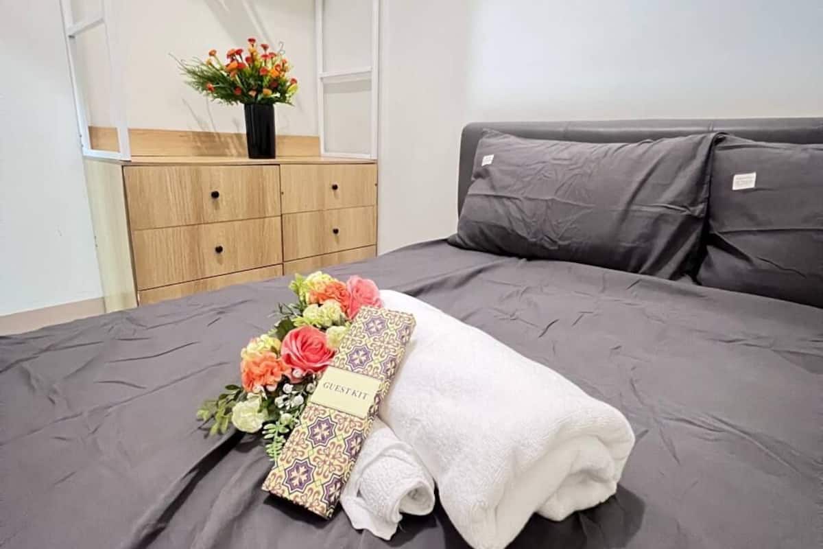 Cama bem arrumada com edredom cinza, toalha branca e arranjo floral com a etiqueta 'GUEST KIT' em um quarto de hotel.