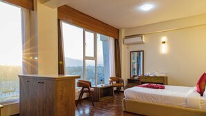 Quarto de hotel com cama, escrivaninha, cadeira, cômoda e uma grande janela com vista para a cidade.
