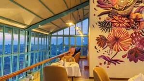 Uma área de refeições com mesas preparadas para dois, grandes janelas com vista para a cidade e um mural de flores tropicais na parede.
