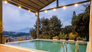Piscina ao ar livre com vista para montanhas e paisagem urbana, cercada por árvores e área coberta com iluminação.