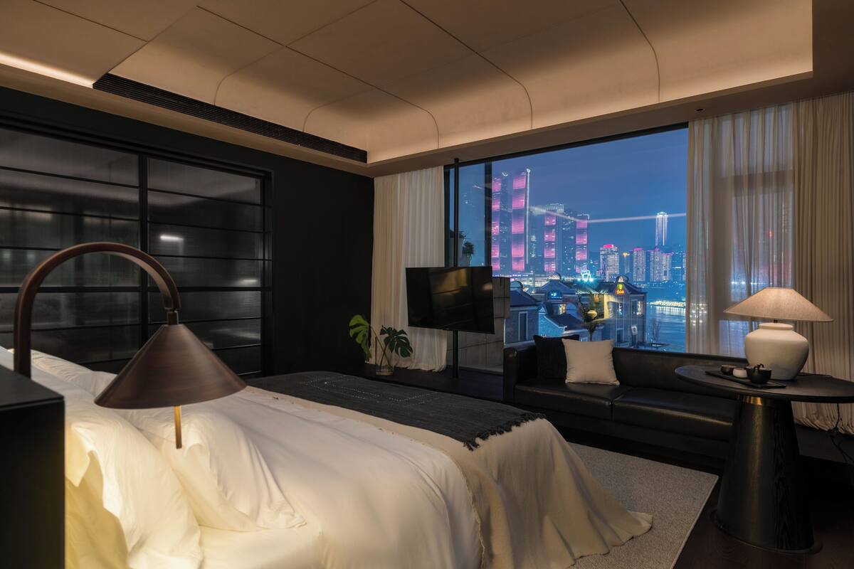 Quarto de hotel moderno com uma cama grande, televisão, sofá e vista para o horizonte urbano à noite.