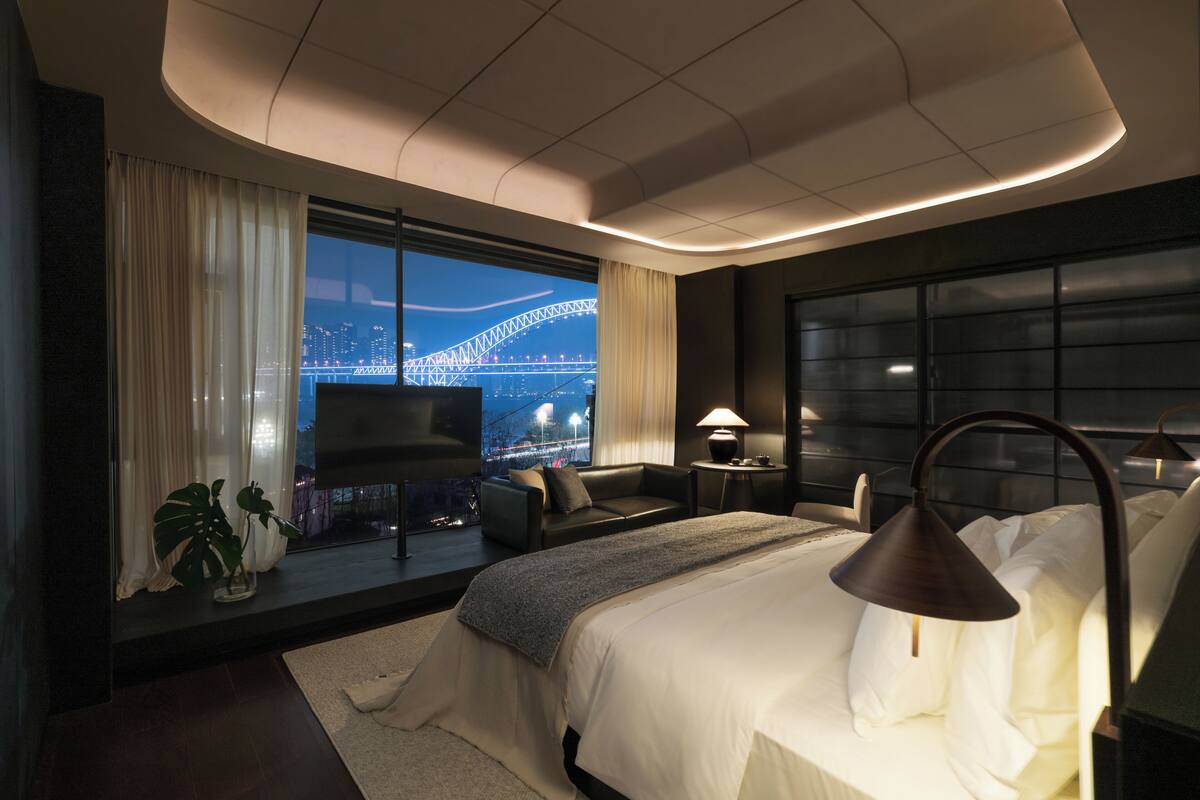 Quarto de hotel moderno com uma cama grande, televisão, sofá e vista para uma ponte à noite.