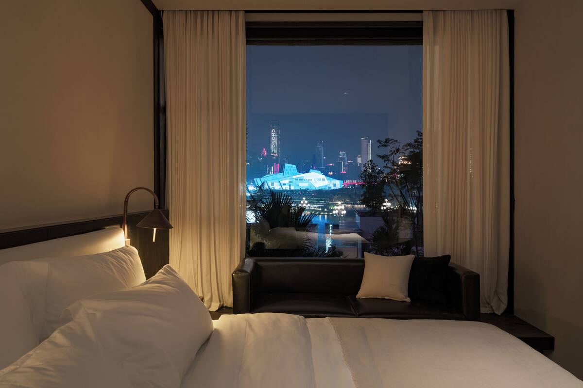 Quarto de hotel com uma cama grande, uma mesa de cabeceira, uma luminária e vista para a paisagem urbana à noite.