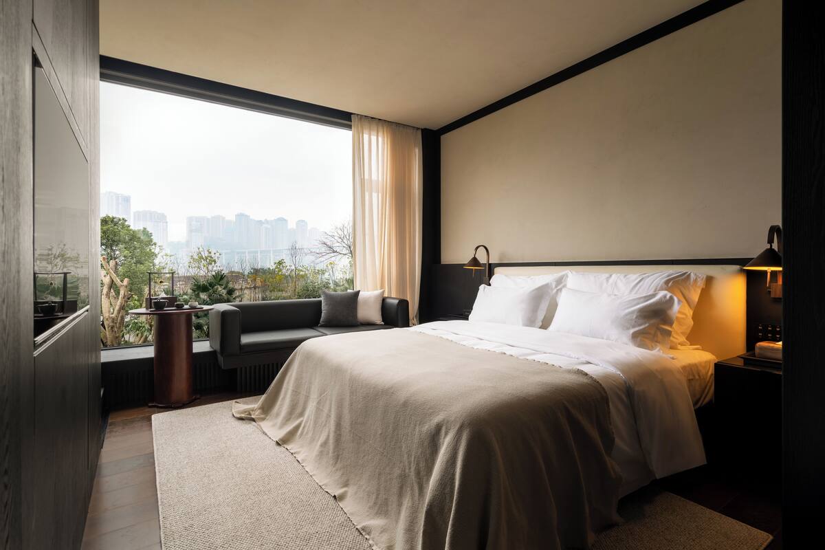 Quarto de hotel moderno com uma cama grande, vista para a cidade, uma mesinha com abajur e um sofá junto à janela.