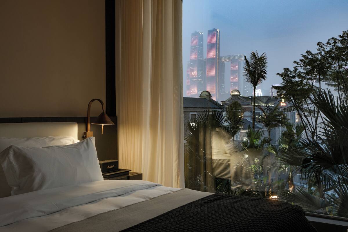 Quarto de hotel com cama, mesa de cabeceira, luminária e vista para edifícios altos e palmeiras pela janela.