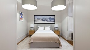Quarto com uma cama grande, duas mesas de cabeceira com abajures, um quadro pendurado na parede com paisagem de montanha e um radiador.