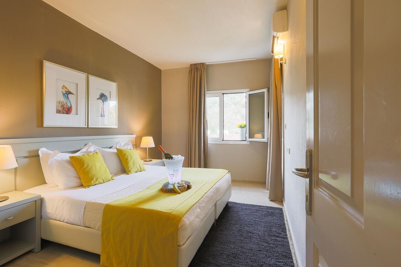 Quarto de hotel com uma cama grande, uma mesa de cabeceira com abajur, uma janela com cortinas e uma pintura na parede.