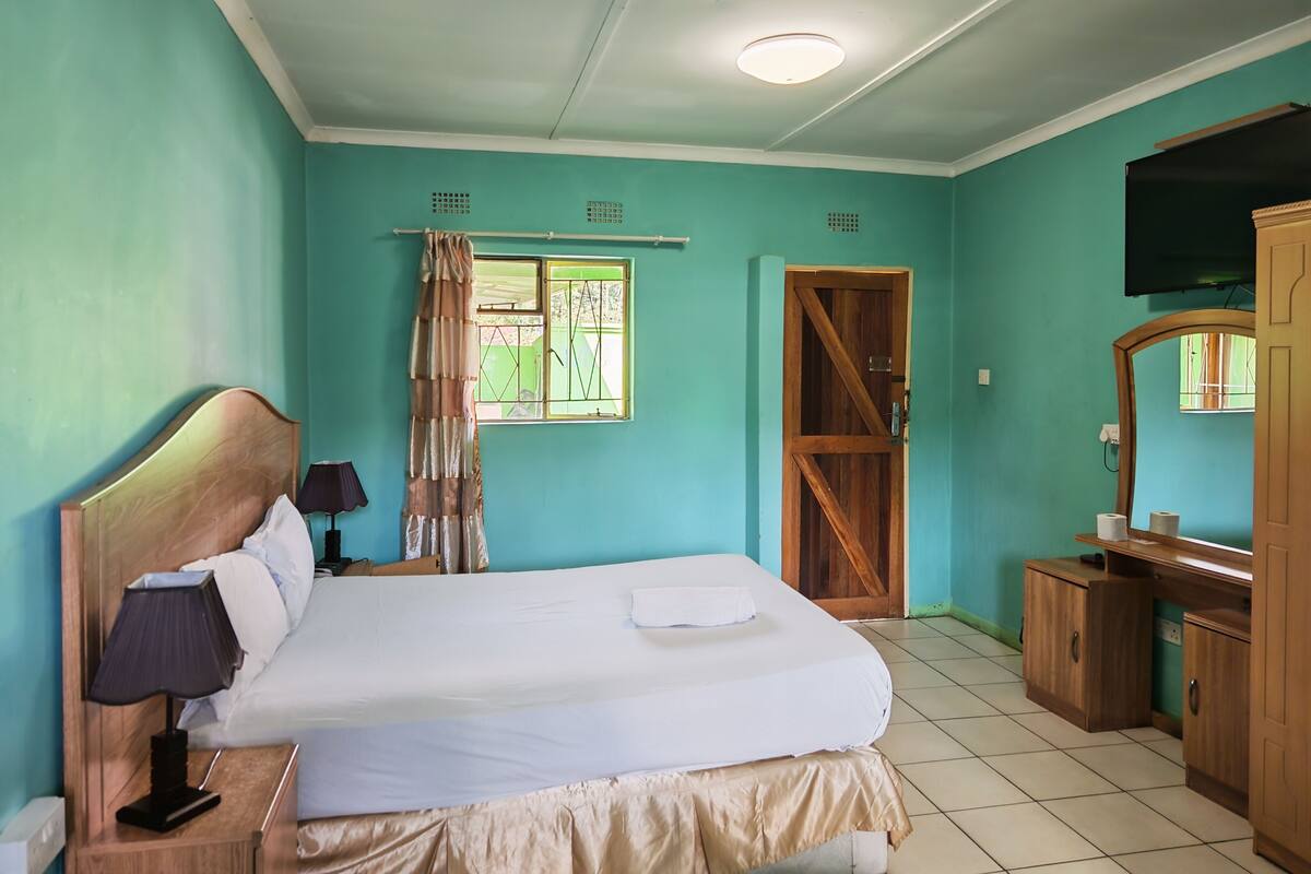 Quarto com cama, mesa de cabeceira, luminária, janela com cortinas, guarda-roupa de madeira e uma televisão.