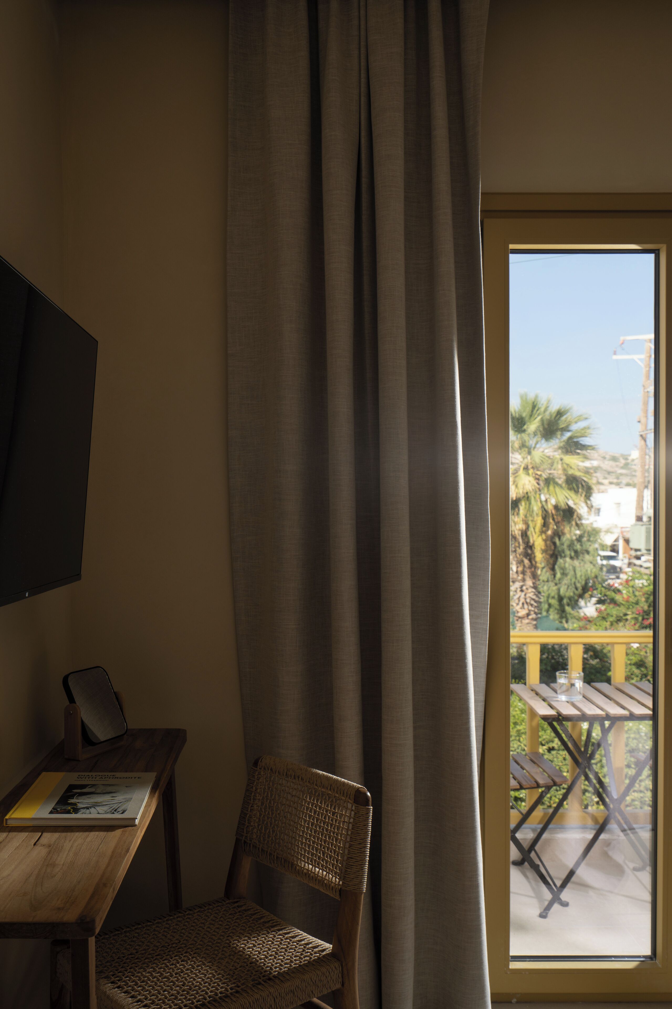 Quarto com escrivaninha de madeira, cadeira de vime, televisão fixada na parede e janela com vista para palmeiras e edifícios.