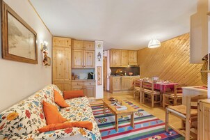 Uma sala de estar aconchegante com sofá, mesa de jantar e área de cozinha com armários de madeira e luminária pendente.