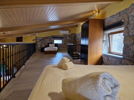 Quarto de hotel com teto de madeira, paredes de pedra, cama com roupa de cama branca, janela com vista e varanda com grade.