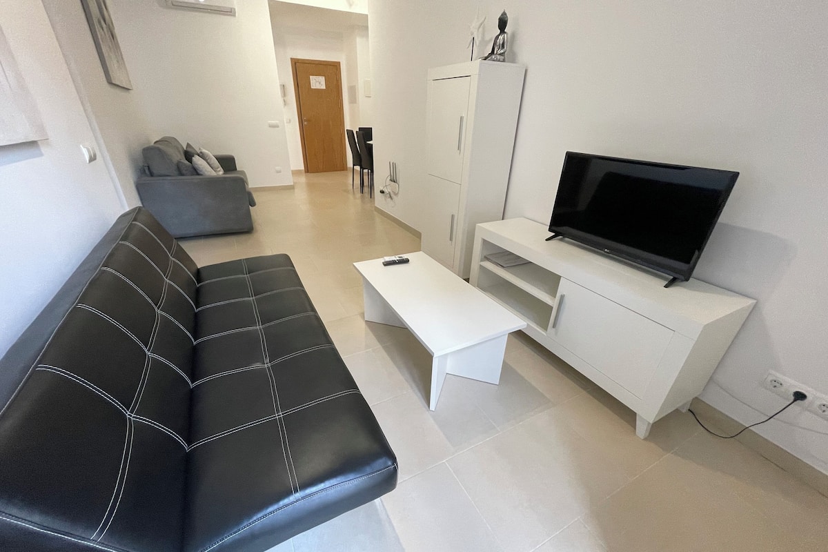 Uma sala de estar moderna com um sofá de couro preto, uma mesa de centro branca, uma TV sobre um móvel branco e um sofá cinzento ao fundo.