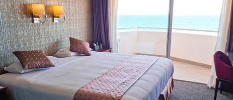 Quarto de hotel com uma cama grande, mesa de cabeceira, escrivaninha e uma cadeira. Há uma janela com vista para o oceano.