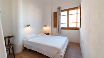 Quarto pequeno e limpo, com uma cama de solteiro, uma cadeira de madeira, mesa de cabeceira e janela com cortinas.