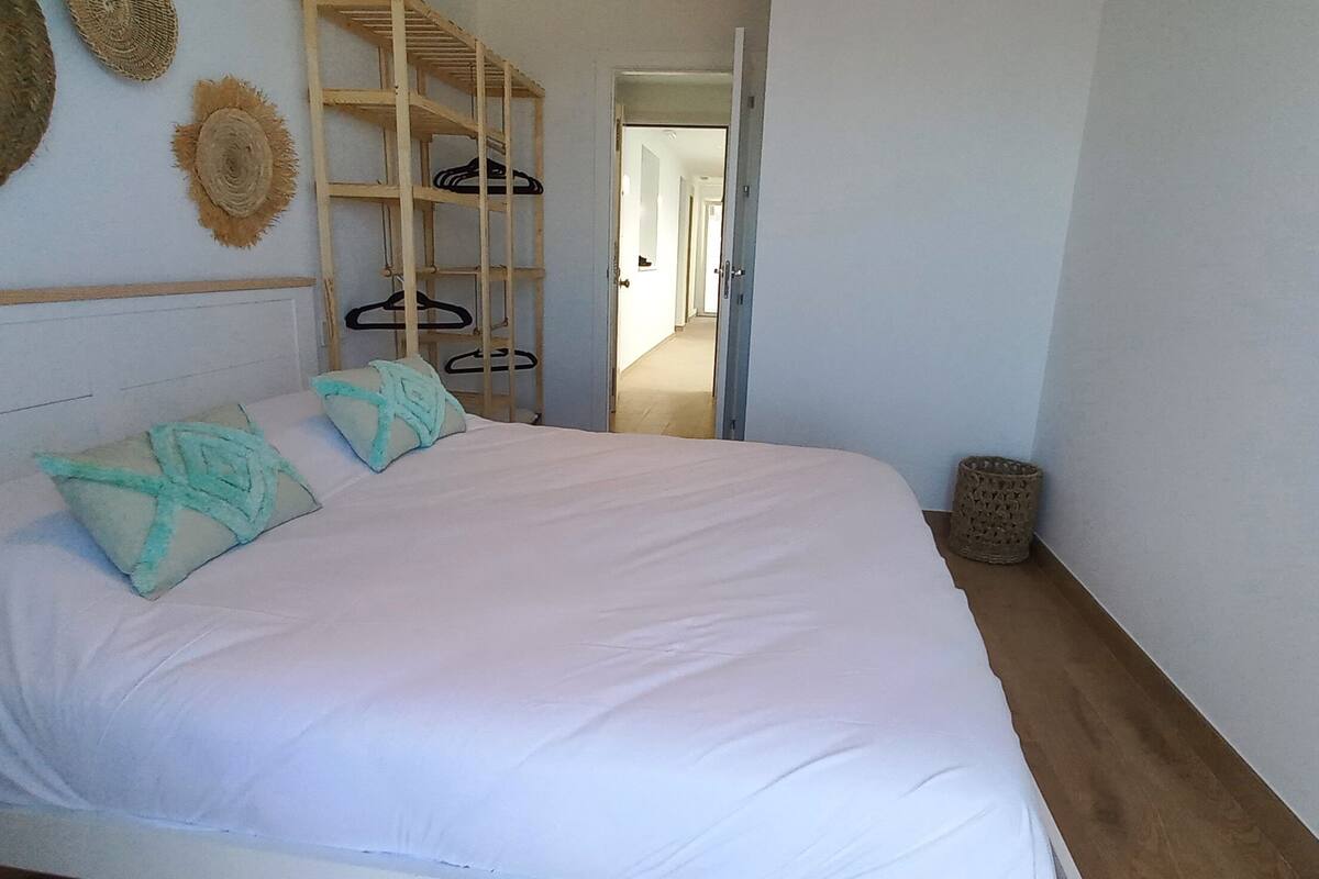 Quarto com cama, almofadas, prateleira de madeira com cabides, cesto de palha e porta que dá para outro cômodo.