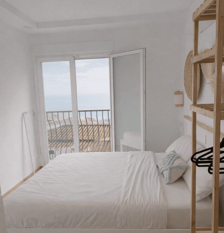 Quarto com cama, prateleira de madeira, varanda e vista para o mar.