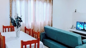 Uma área de refeições com mesa, cadeiras e um vaso de flores. Há um sofá e uma televisão ao fundo.