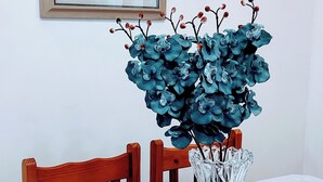 Uma mesa de jantar com um vaso de vidro transparente com flores azuis, cadeiras de madeira e uma obra de arte emoldurada na parede.