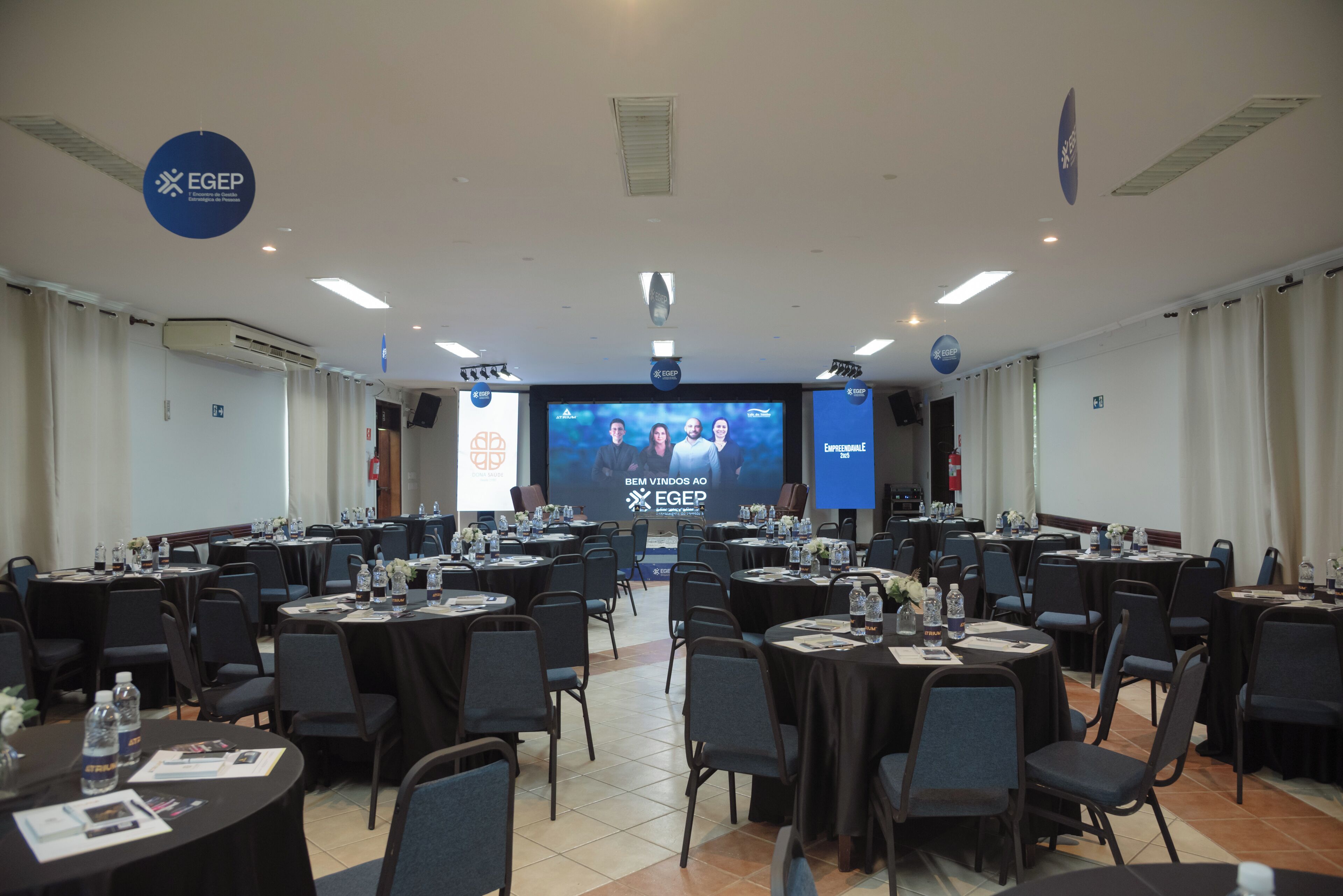 Uma sala de conferências com várias mesas, cadeiras e uma tela grande exibindo uma apresentação.