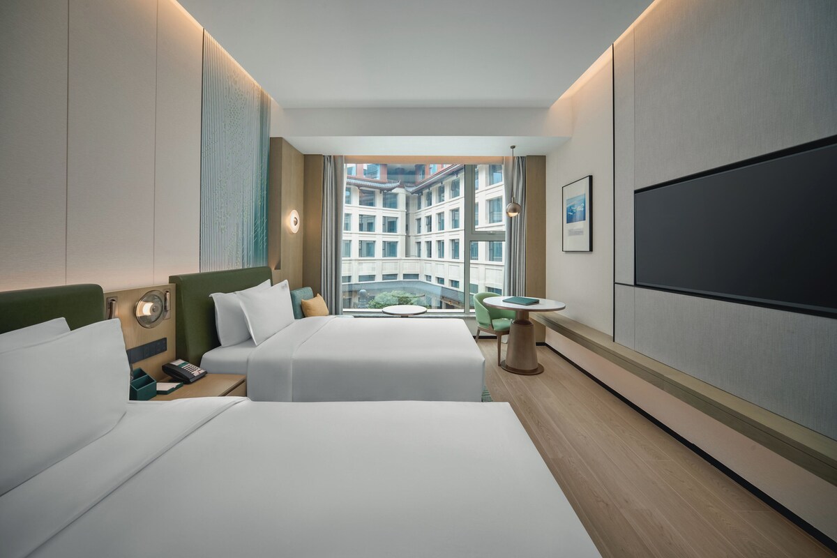 Quarto de hotel moderno com duas camas, uma mesinha, uma cadeira e uma televisão grande.