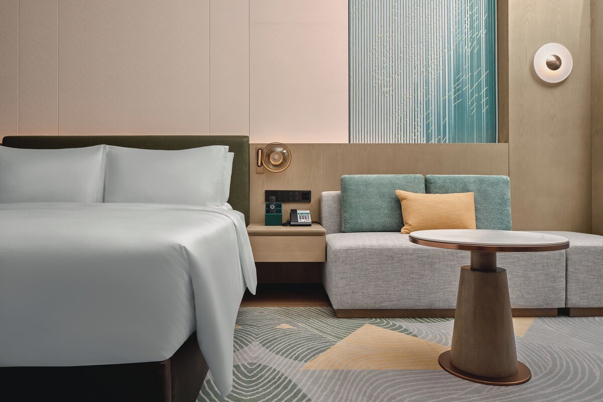 Quarto de hotel moderno com cama, sofá, mesa pequena e luminária fixada na parede.