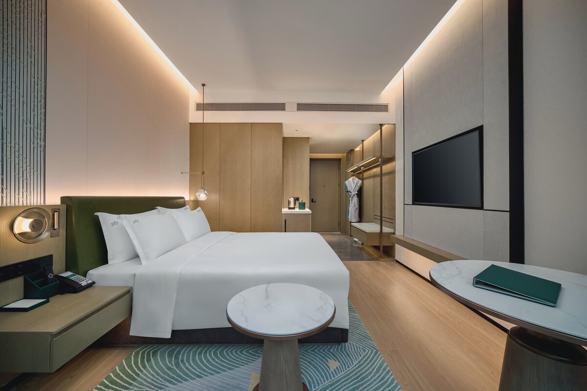 Quarto de hotel moderno com cama, mesa de cabeceira, televisão e uma pequena mesa redonda.