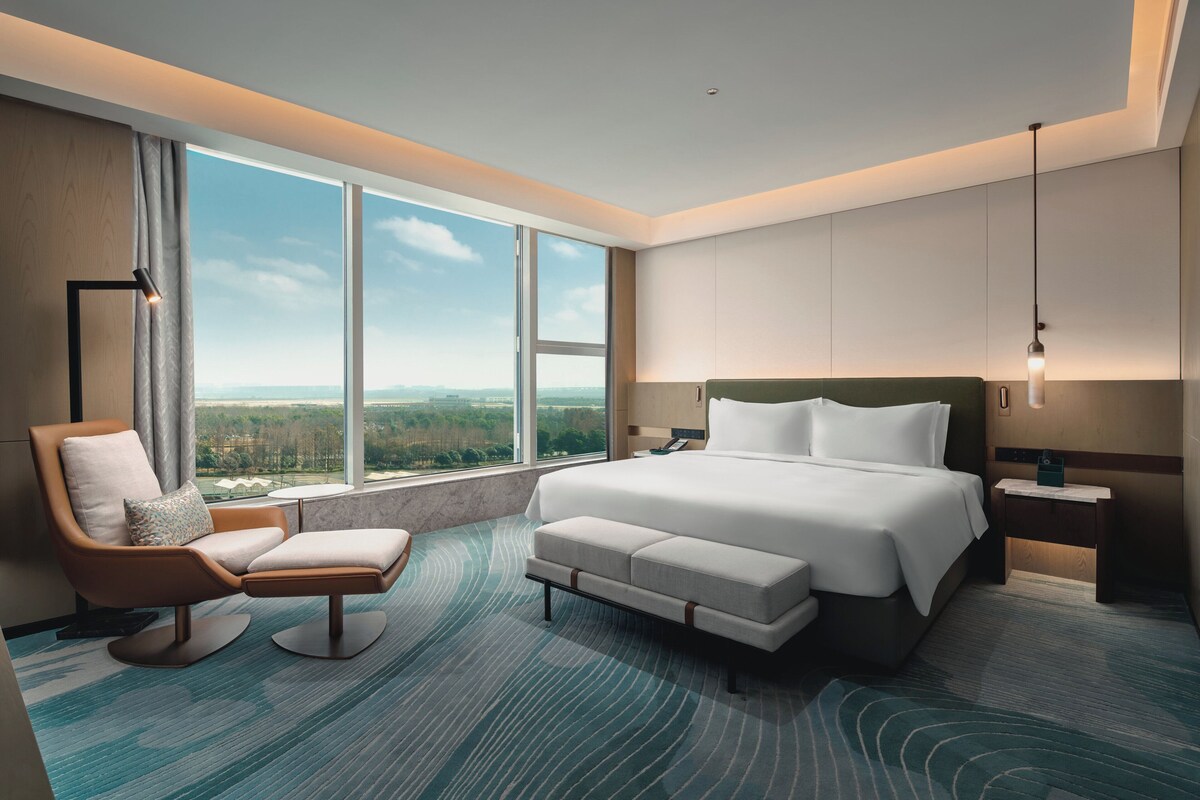 Quarto de hotel moderno com uma cama grande, área de estar, escrivaninha e vista para o exterior.