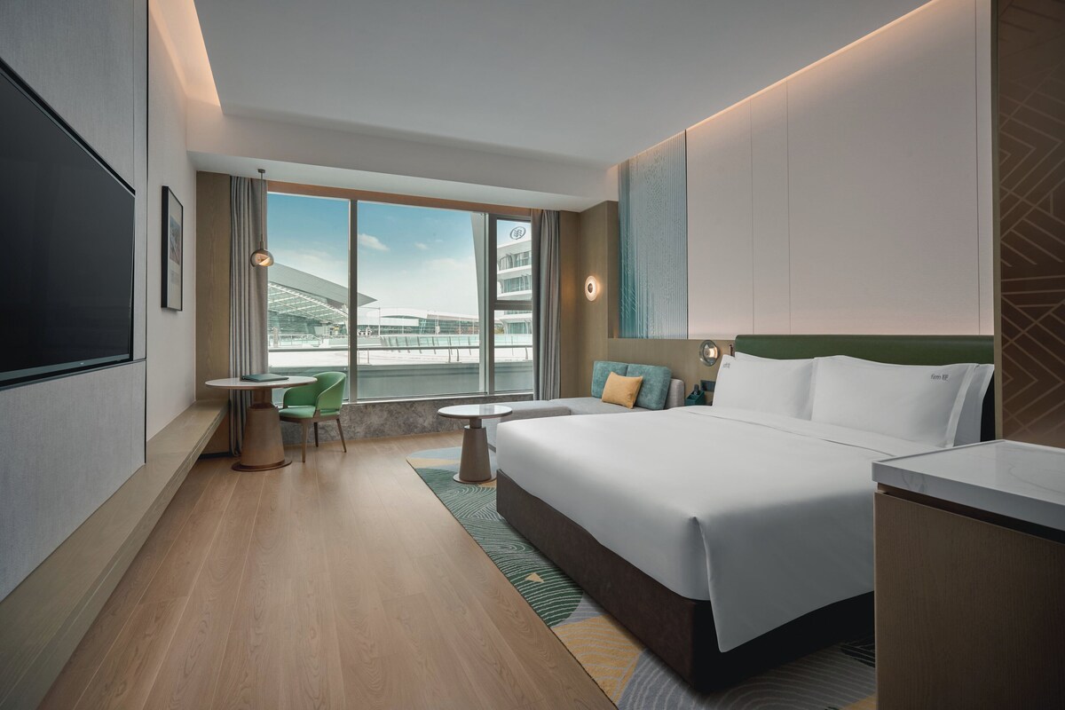 Quarto de hotel moderno com uma cama grande, uma mesinha, uma cadeira e uma televisão.